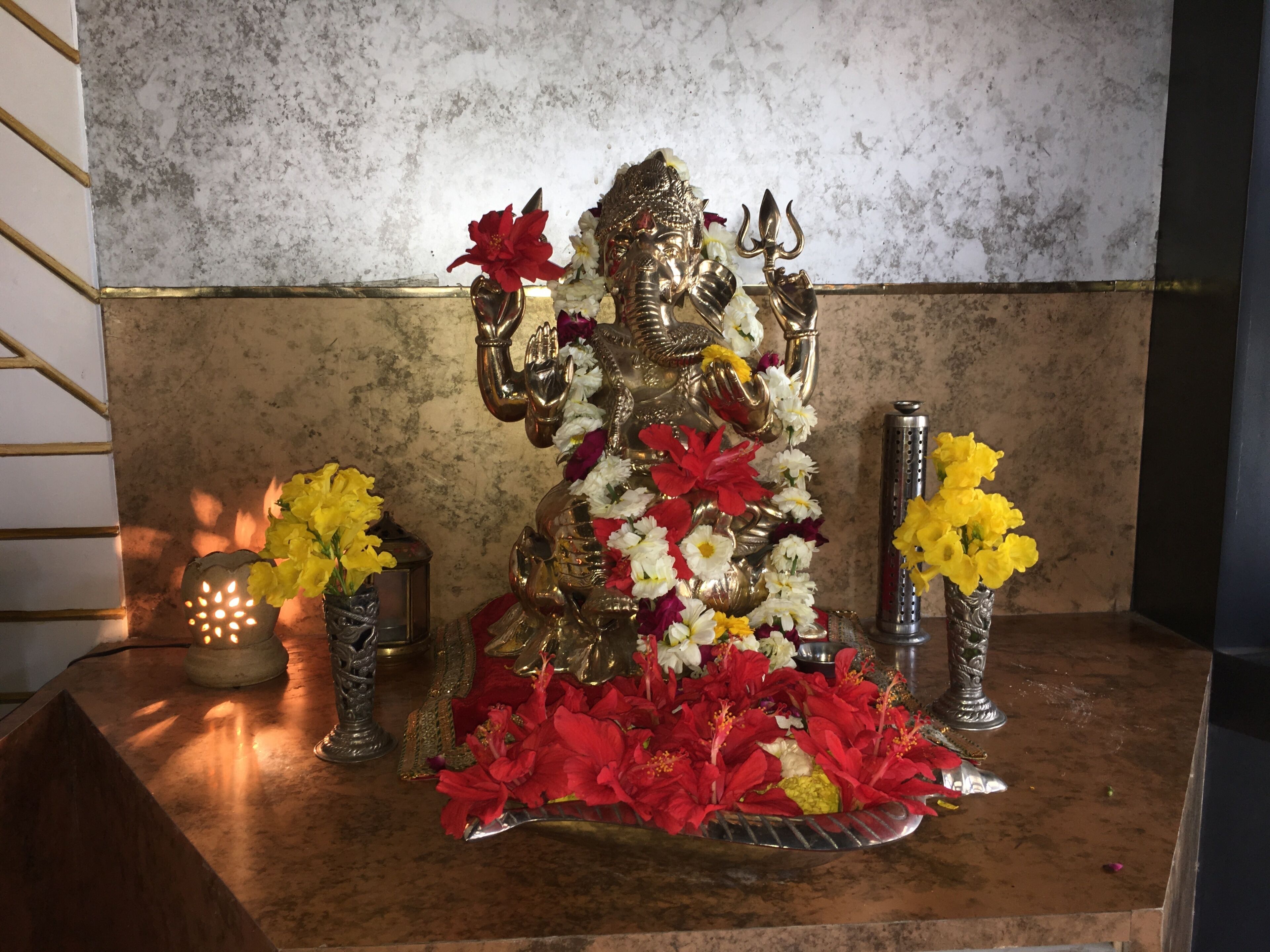 Uma estátua de uma divindade adornada com flores e um tridente, colocada sobre um altar de mármore com castiçais e vasos de flores.