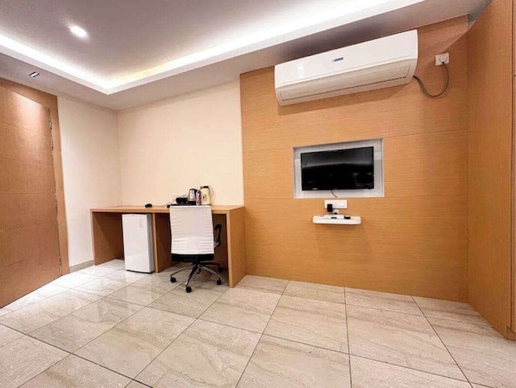 Quarto pequeno e bem iluminado, com uma mesa, televisão fixada na parede e aparelho de ar condicionado.