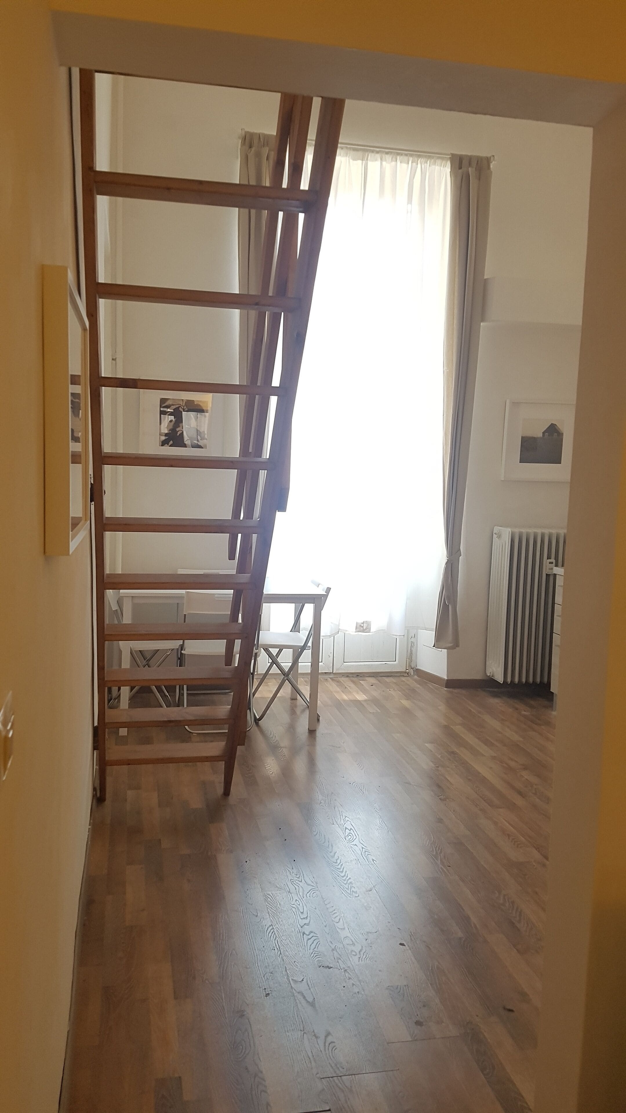 Quarto com escada de madeira, radiador e janela com cortinas.