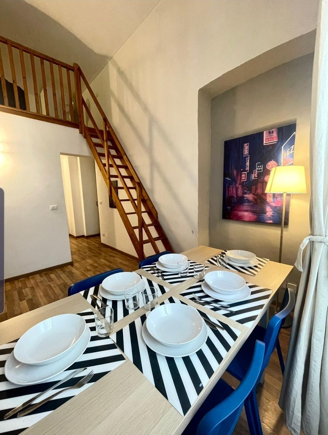 Uma sala de jantar com uma mesa posta para quatro pessoas, uma escadaria e um quadro com vista para a cidade.