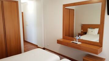 Quarto de hotel com estrutura de cama de madeira, mesa de cabeceira com abajur, espelho grande e um pequeno banquinho de madeira.