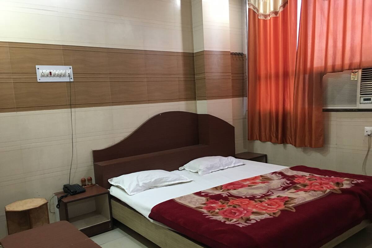 Quarto de hotel com cama de cabeceira em madeira, mesa de cabeceira com abajur, pequena escrivaninha e colcha vermelha com padrão floral.