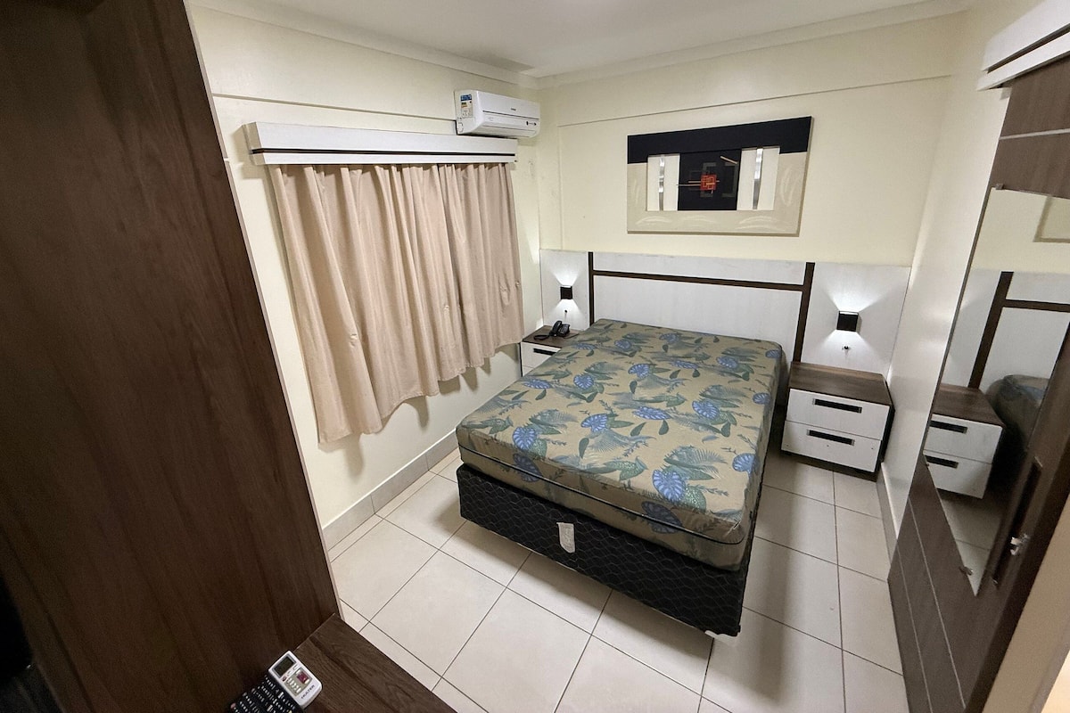 Quarto com cama, mesas de cabeceira, ar condicionado, espelho e cortina.