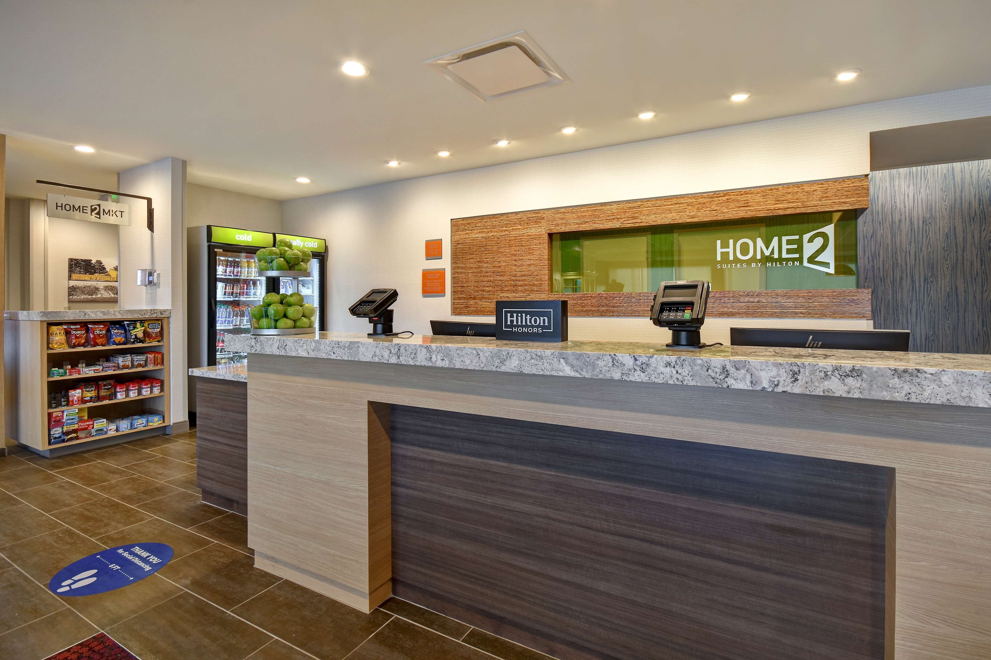 Uma área de recepção de hotel com balcão de mármore, exposição de frutas e placa com 'Home 2 Suites by Hilton'.