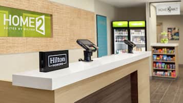 Uma receção de hotel com um quiosque digital, um letreiro 'HOME 2 Suites by Hilton' e um sinal de 'Hilton Honors'.