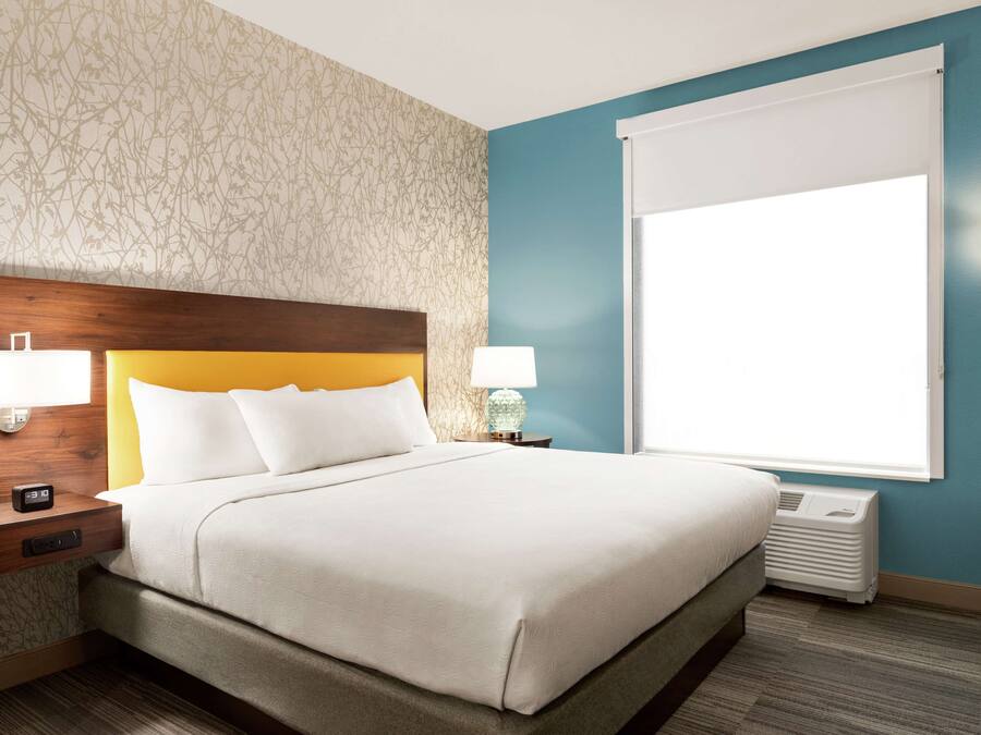 Quarto de hotel com uma cama grande, uma mesa de cabeceira com luminária, um aquecedor fixado na parede e uma janela com persianas.