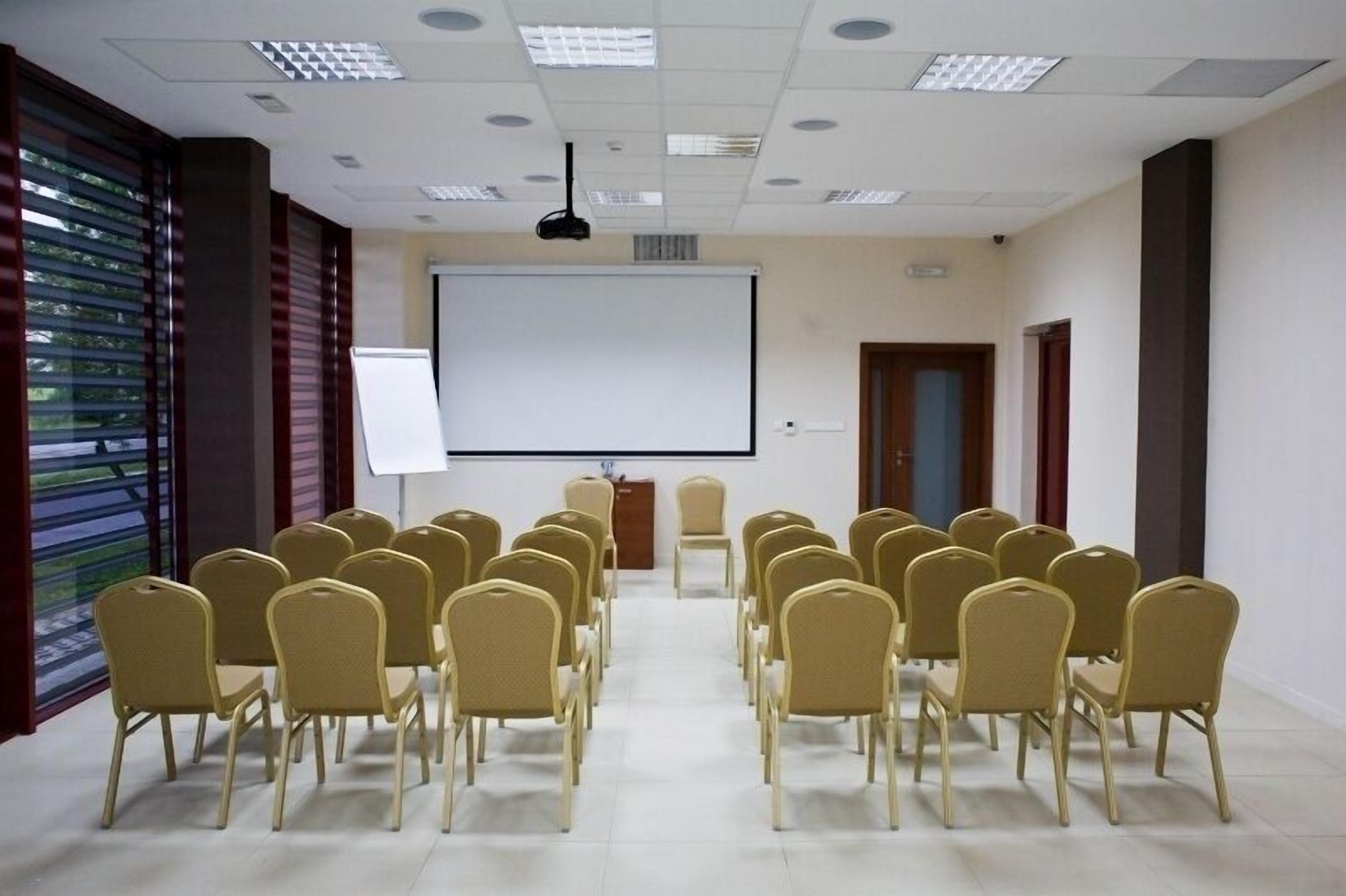 Uma sala de conferências com fileiras de cadeiras voltadas para uma tela branca grande, um projetor e uma pequena mesa com uma garrafa de água.