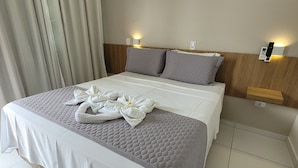 Quarto de hotel com uma cama grande, duas almofadas, uma mesa de cabeceira com abajur e uma cortina.