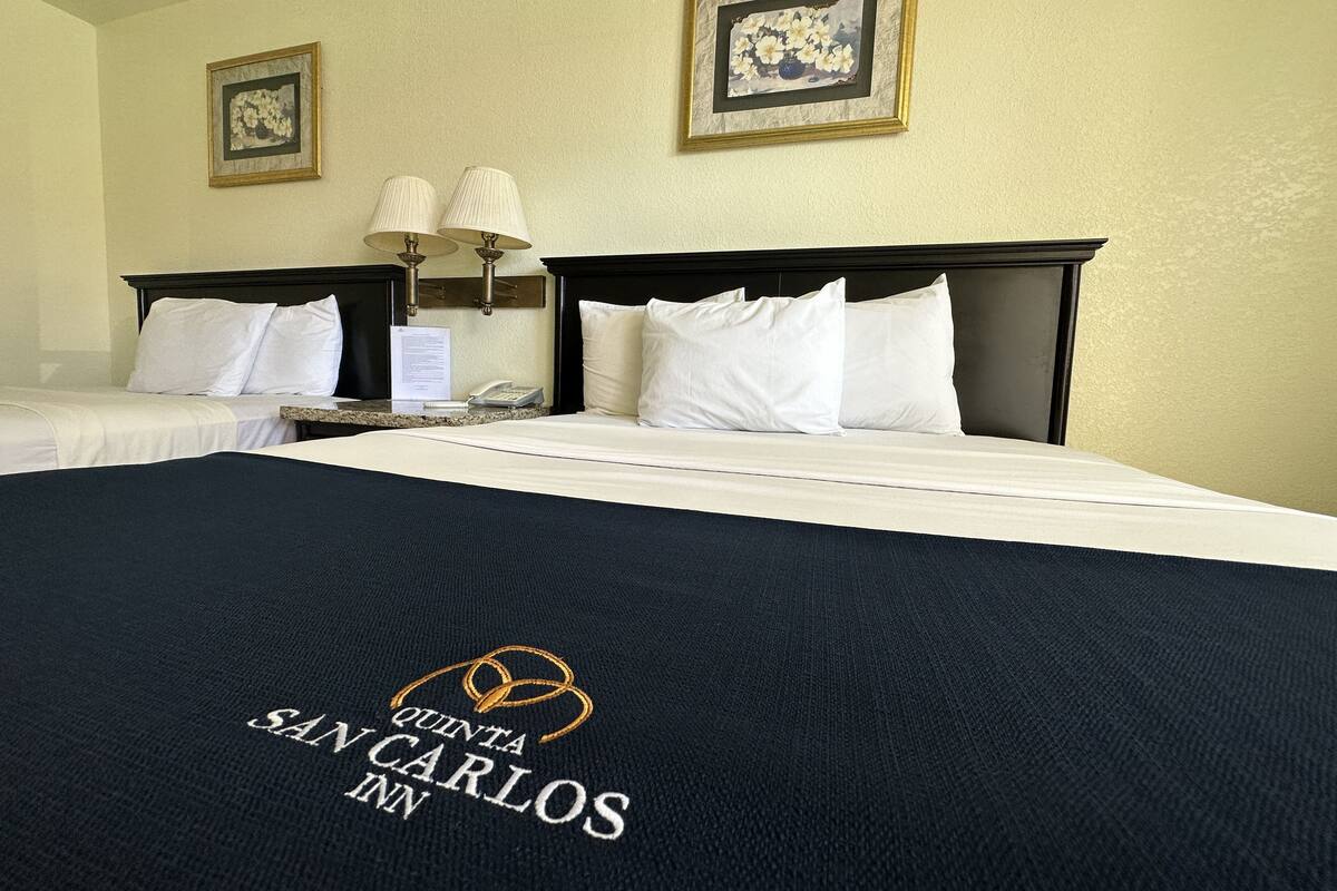 Quarto de hotel com duas camas, mesa de cabeceira, luminária e quadros na parede.
