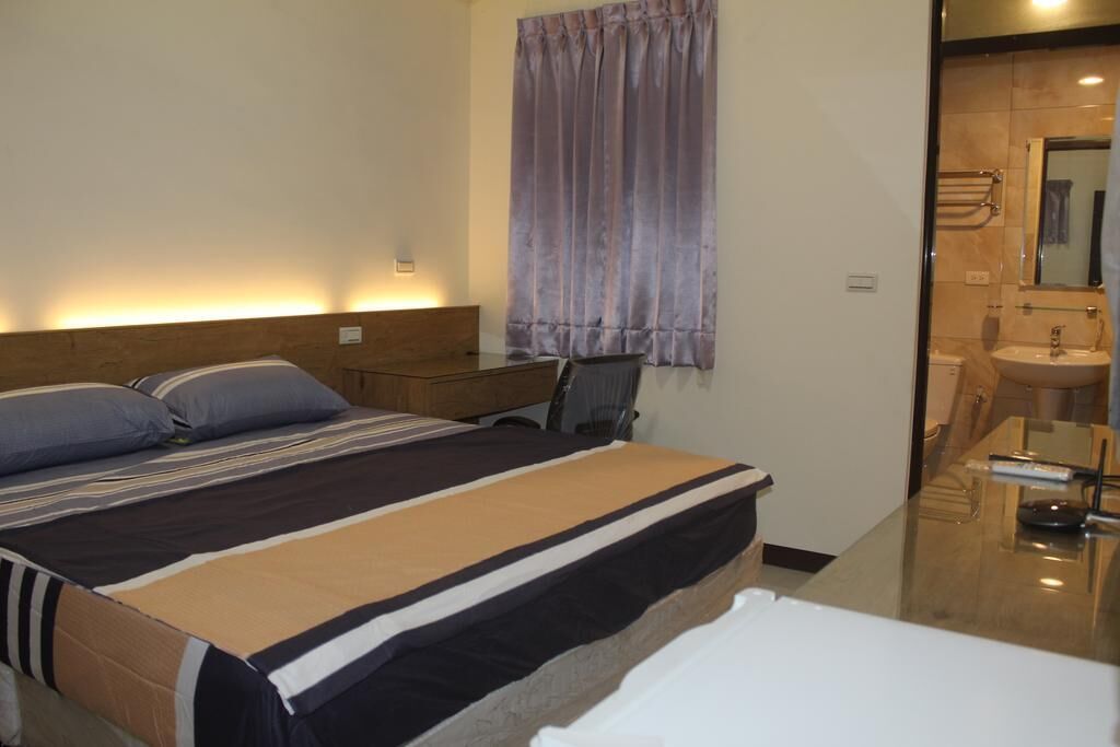 Quarto de hotel com cama, escrivaninha, cadeira, janela com cortinas e banheiro visível por uma porta aberta.