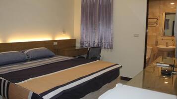 Quarto de hotel com cama, escrivaninha, cadeira, janela com cortinas e banheiro visível por uma porta aberta.