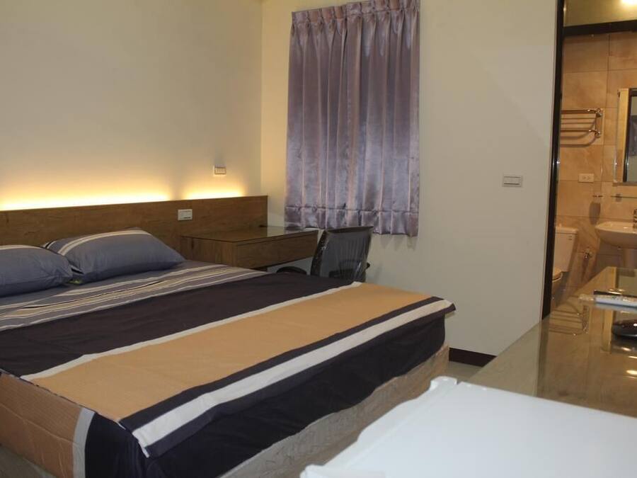 Quarto de hotel com cama, escrivaninha, cadeira, janela com cortinas e banheiro visível por uma porta aberta.