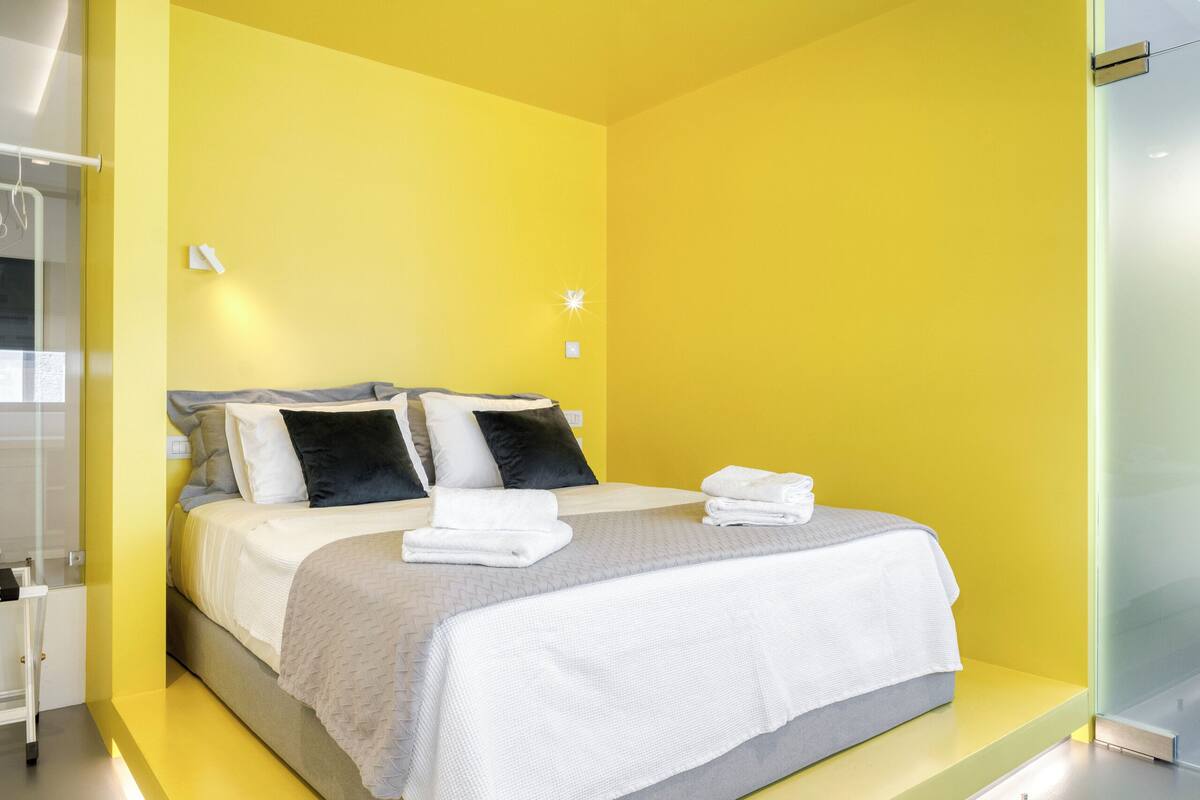 Um quarto moderno com parede amarela de destaque, cama bem arrumada com roupa de cama branca e box de chuveiro de vidro.