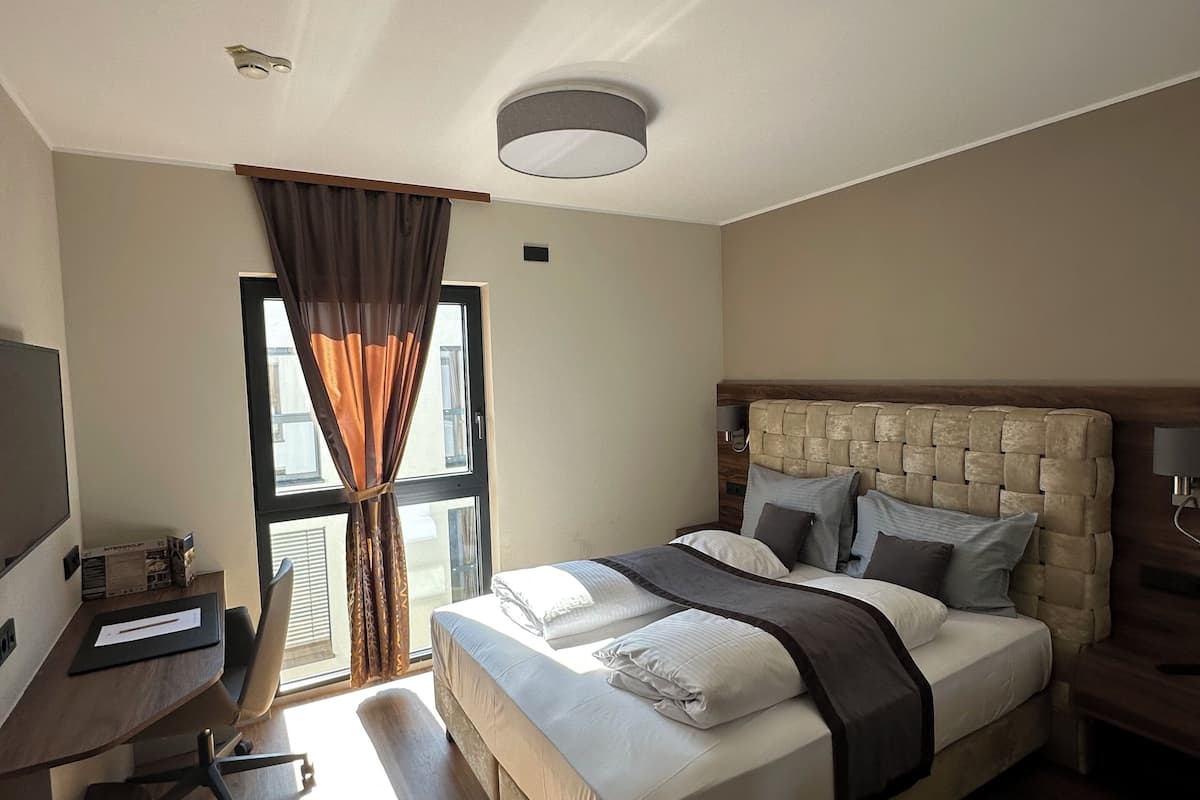 Quarto de hotel com cama, escrivaninha, cadeira e janela com cortinas.