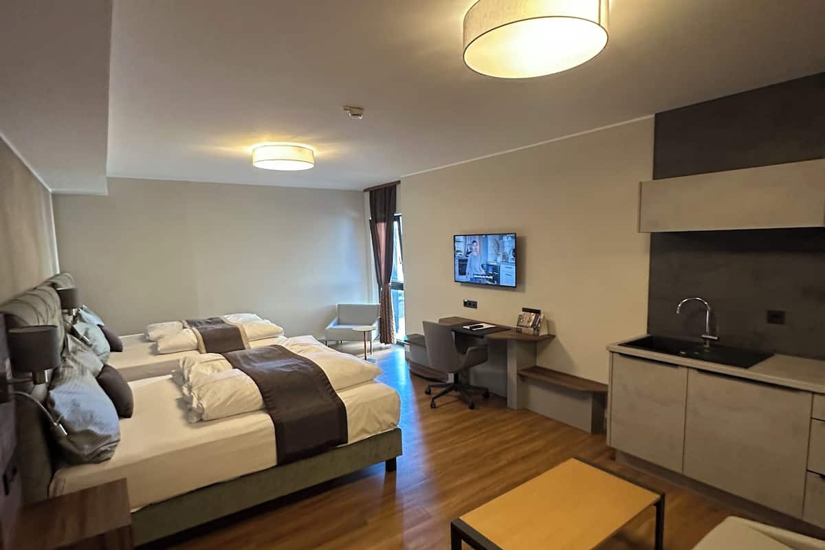 Quarto de hotel moderno com cama, escrivaninha, cozinha compacta e televisão.
