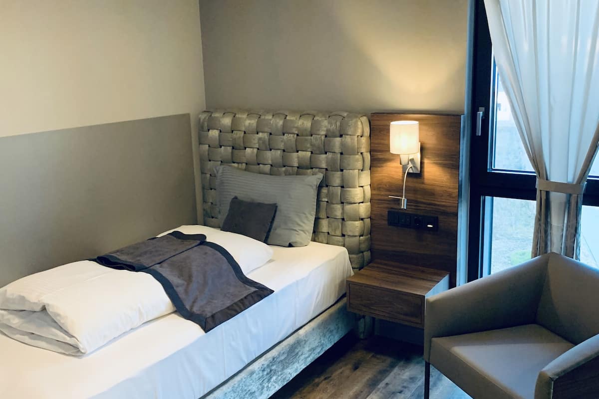 Quarto de hotel com cama, cadeira, mesa de cabeceira com luminária e janela com cortinas.