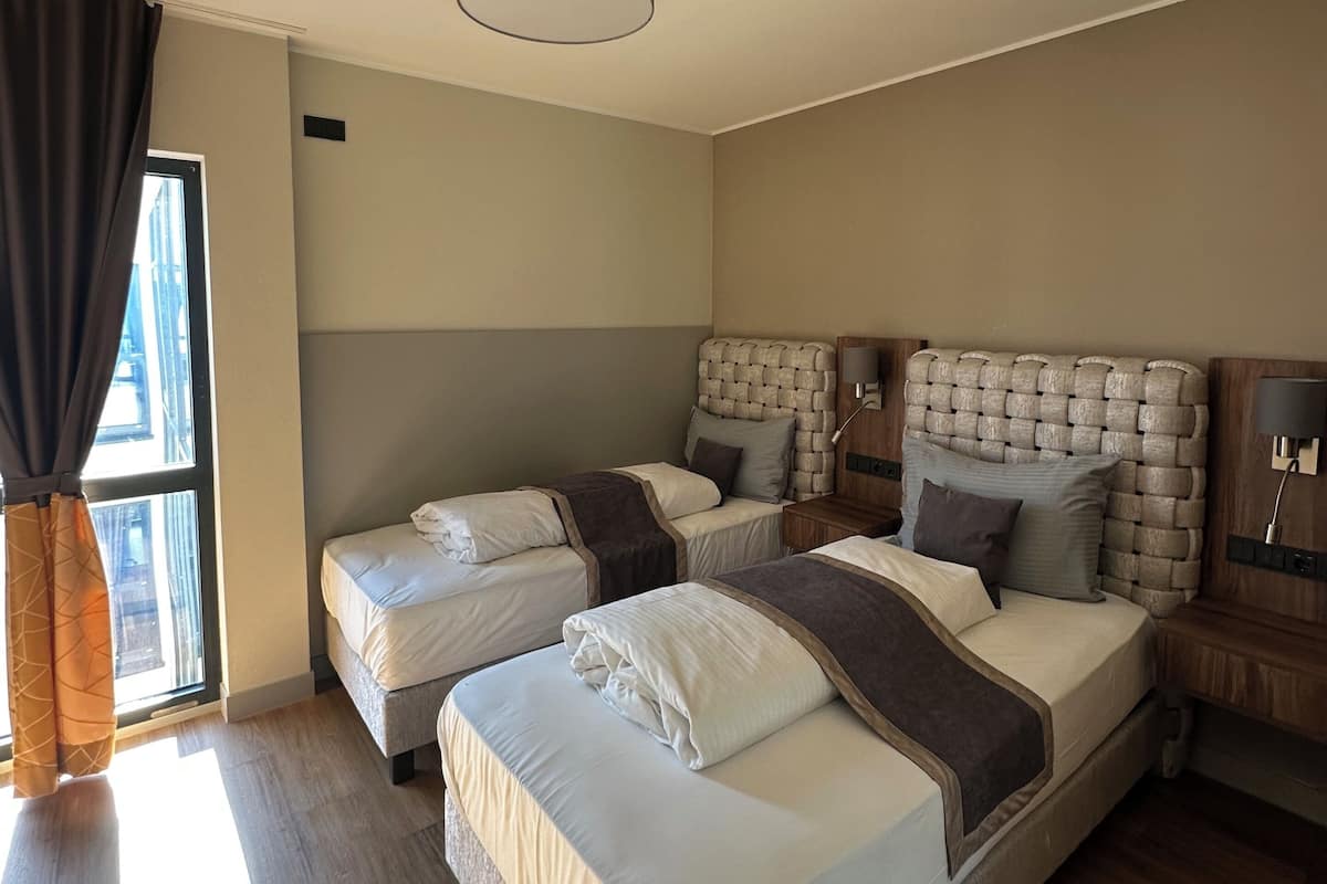 Quarto de hotel com duas camas, uma janela grande e uma luminária de teto moderna.
