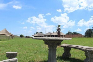 Um campo gramado com um banco de pedra e uma mesa de pedra em primeiro plano, e um edifício com telhado de palha ao fundo.