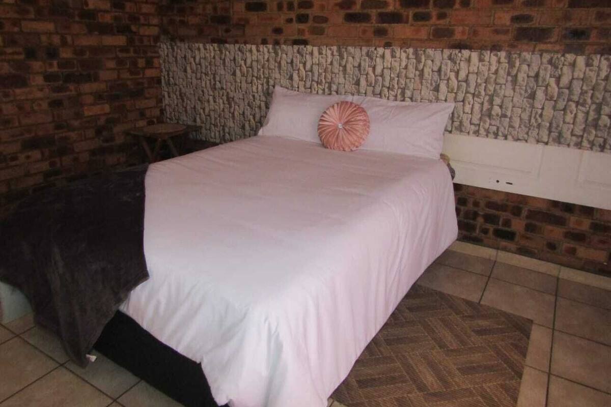 Quarto com parede de tijolos, cabeceira de pedra, cama com colcha branca e tapete estampado.