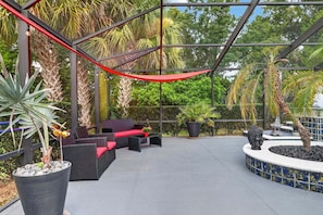Um pátio externo com mobiliário de vime, um toldo vermelho e plantas tropicais.
