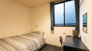 Quarto pequeno e bem organizado, com uma cama de solteiro, uma escrivaninha com computador e uma janela com cortinas.