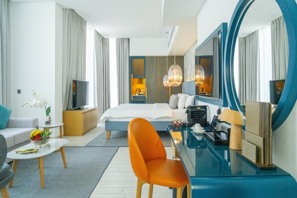 Quarto de hotel moderno com cama, sofá, mesa de jantar com cadeiras, televisão e minibar.