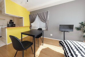 Quarto compacto com cama, televisão, mesa de jantar com cadeiras, micro-ondas e pequena cozinha.