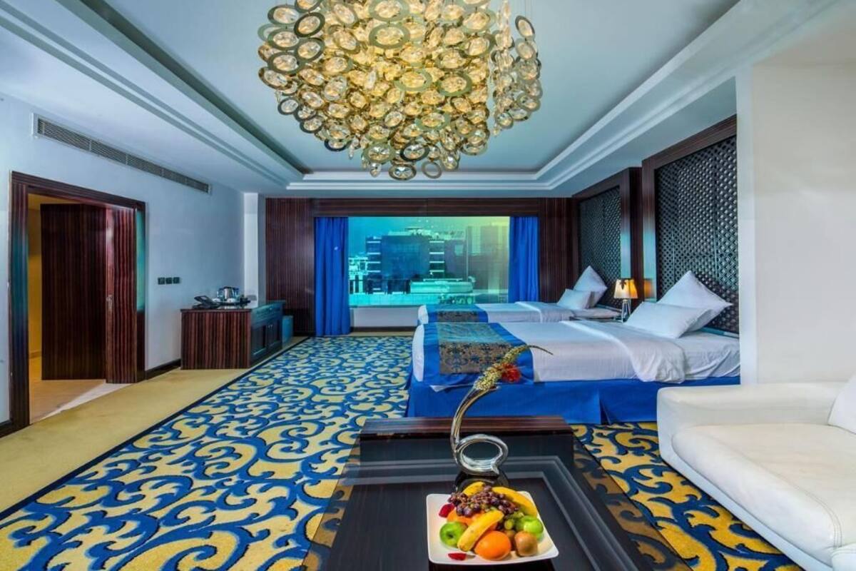 Quarto de hotel com um candelabro grande, uma cama com colcha azul, um sofá, uma mesa de centro com frutas e vista para a paisagem urbana.