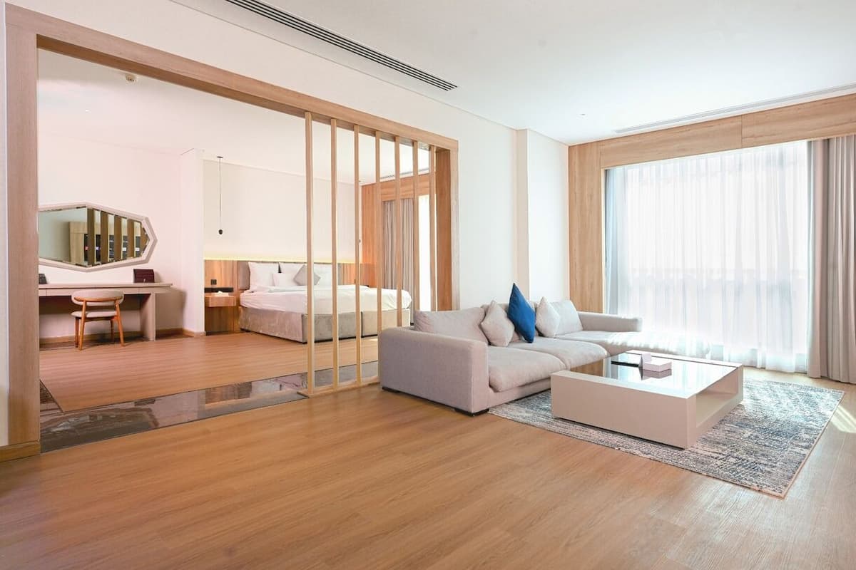 Quarto de hotel moderno com chão de madeira, sofá, cama, escrivaninha e uma janela grande com cortinas.
