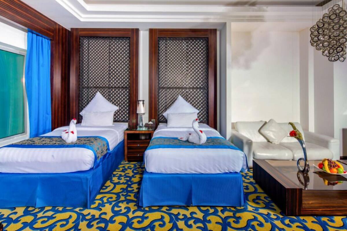 Quarto de hotel com duas camas, um sofá, uma mesa de centro com frutas e uma peça decorativa na parede.