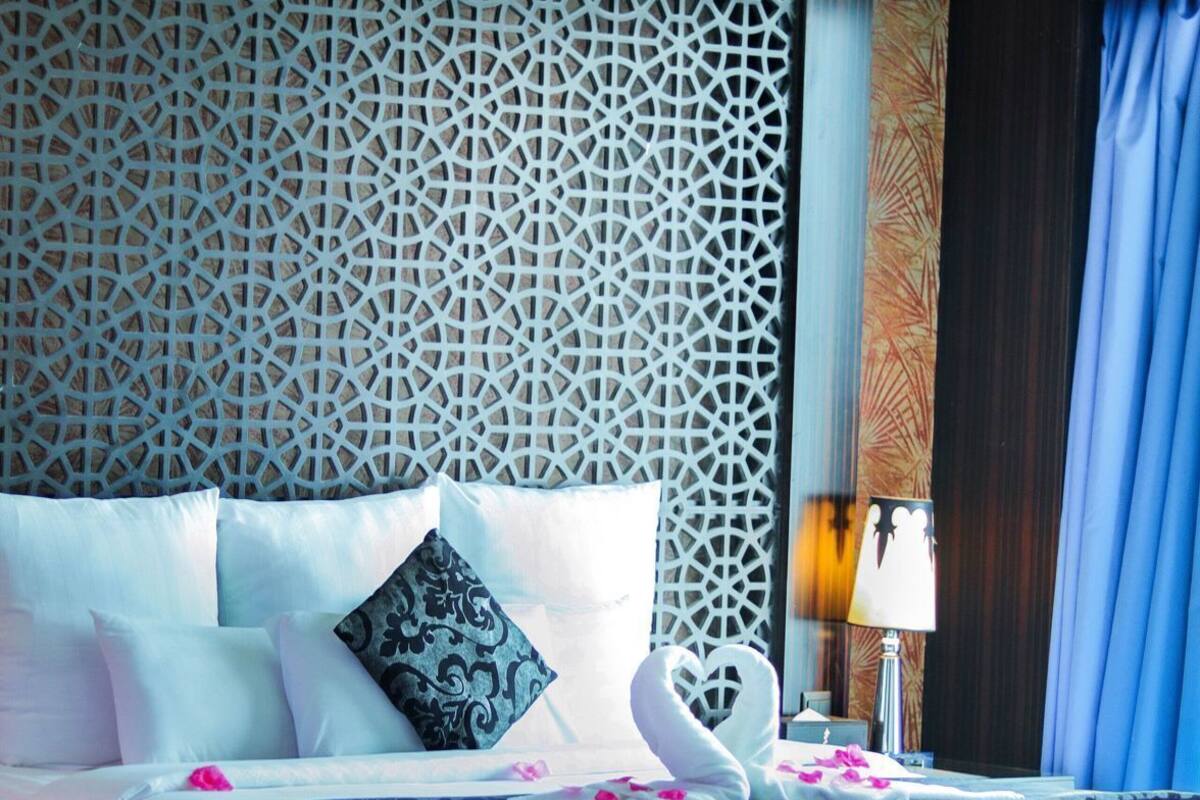 Quarto de hotel com cama, parede decorativa, cortinas e mesa de cabeceira com abajur.