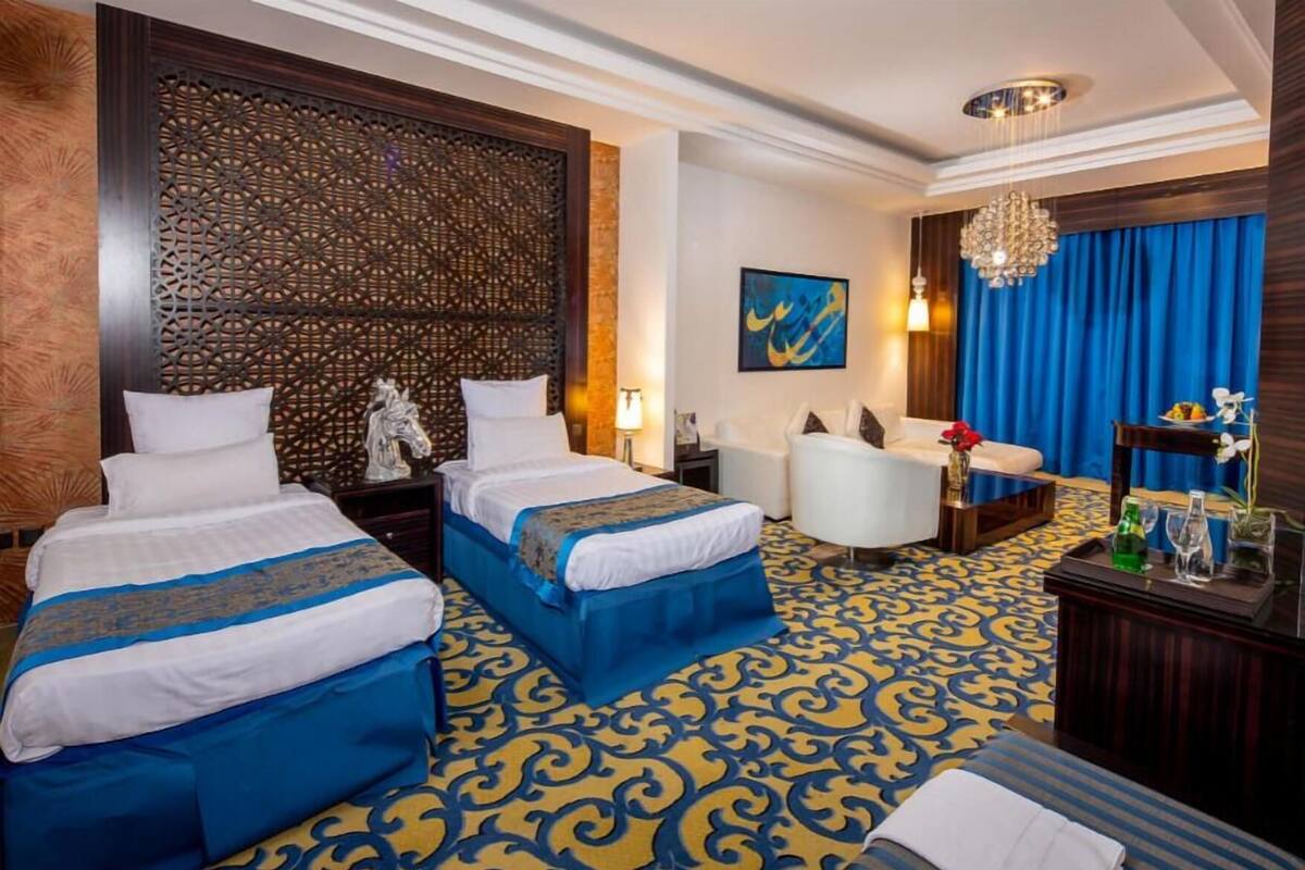 Quarto de hotel com duas camas, um sofá, uma escrivaninha, um candelabro e um painel decorativo na parede.