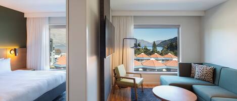Quarto de hotel com cama, sofá, poltrona, mesinha e uma grande janela com vista para edifícios e montanhas.