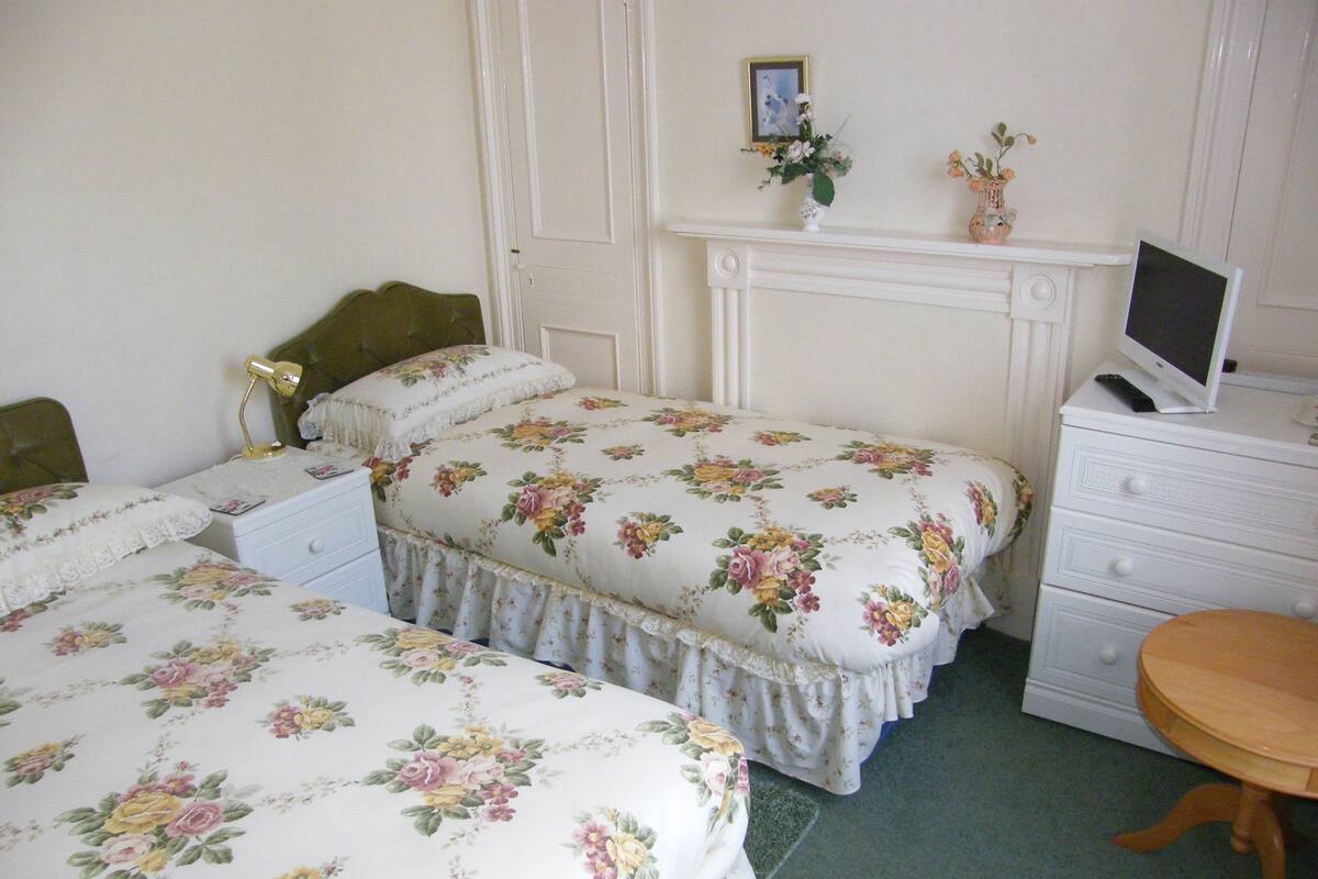 Quarto com duas camas de solteiro, cômoda, uma mesinha e televisão sobre um móvel.