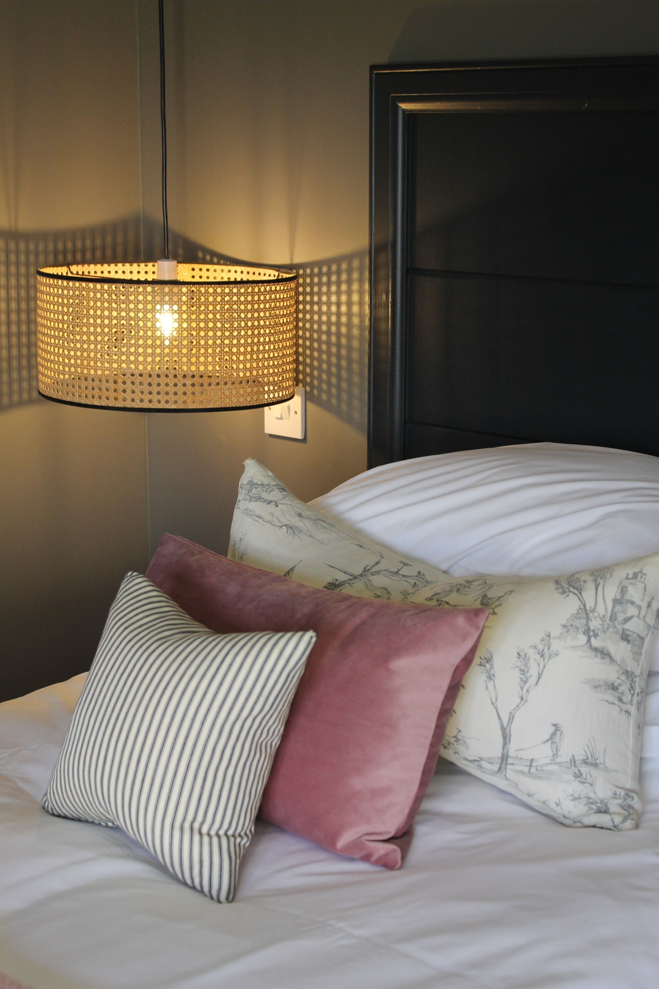 Uma cama bem arrumada com lençol branco e vários travesseiros, incluindo listras e estampas florais. Uma luminária trançada pendurada sobre a cama, proporcionando iluminação suave.