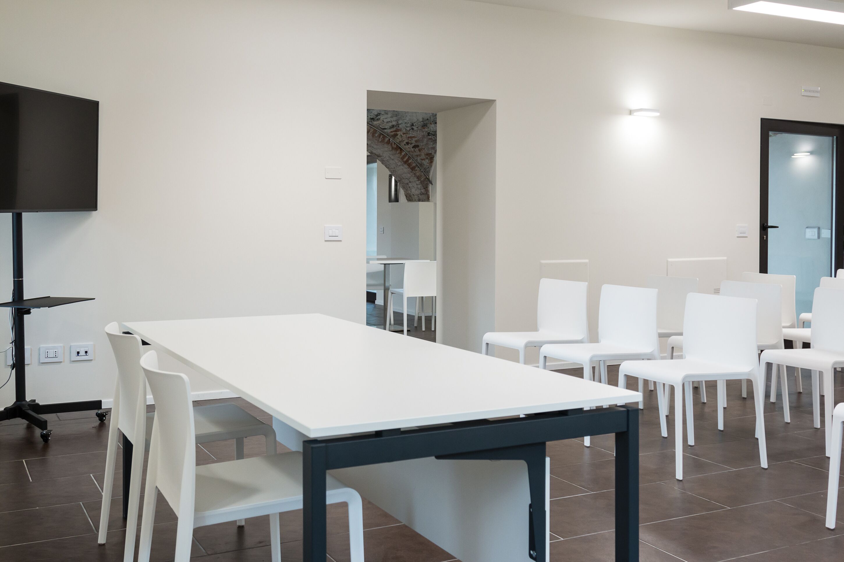 Uma sala de conferências com mesa e cadeiras brancas, uma TV de tela plana e uma porta com painel de vidro.