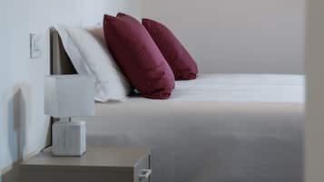 Cama bem arrumada com almofadas brancas e bordô, mesa de cabeceira com abajur e interruptor de luz fixado na parede.