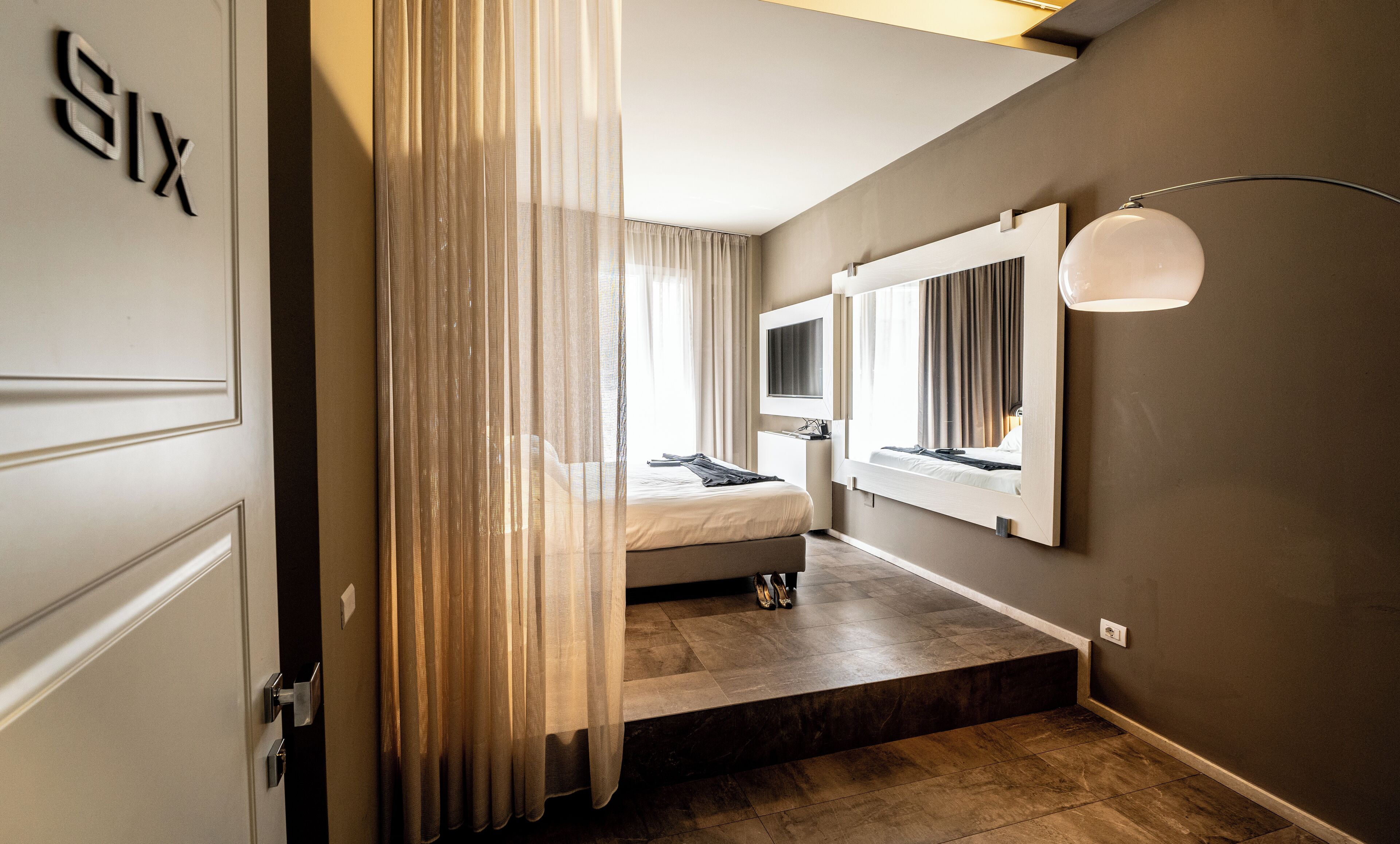 Quarto de hotel moderno com uma cama, um espelho grande, uma televisão e uma porta com a placa 'SIX'.