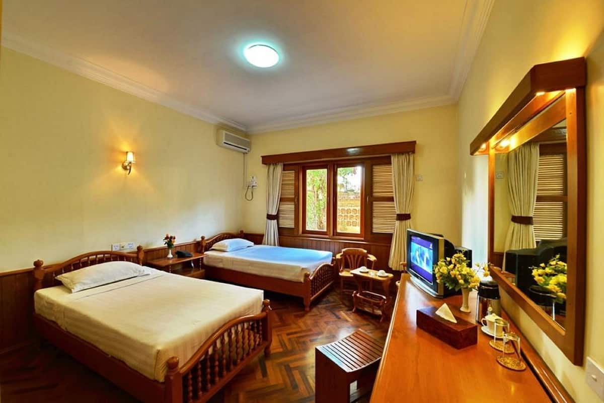Quarto de hotel com duas camas de solteiro, uma escrivaninha com televisão, um espelho e uma janela com cortinas.