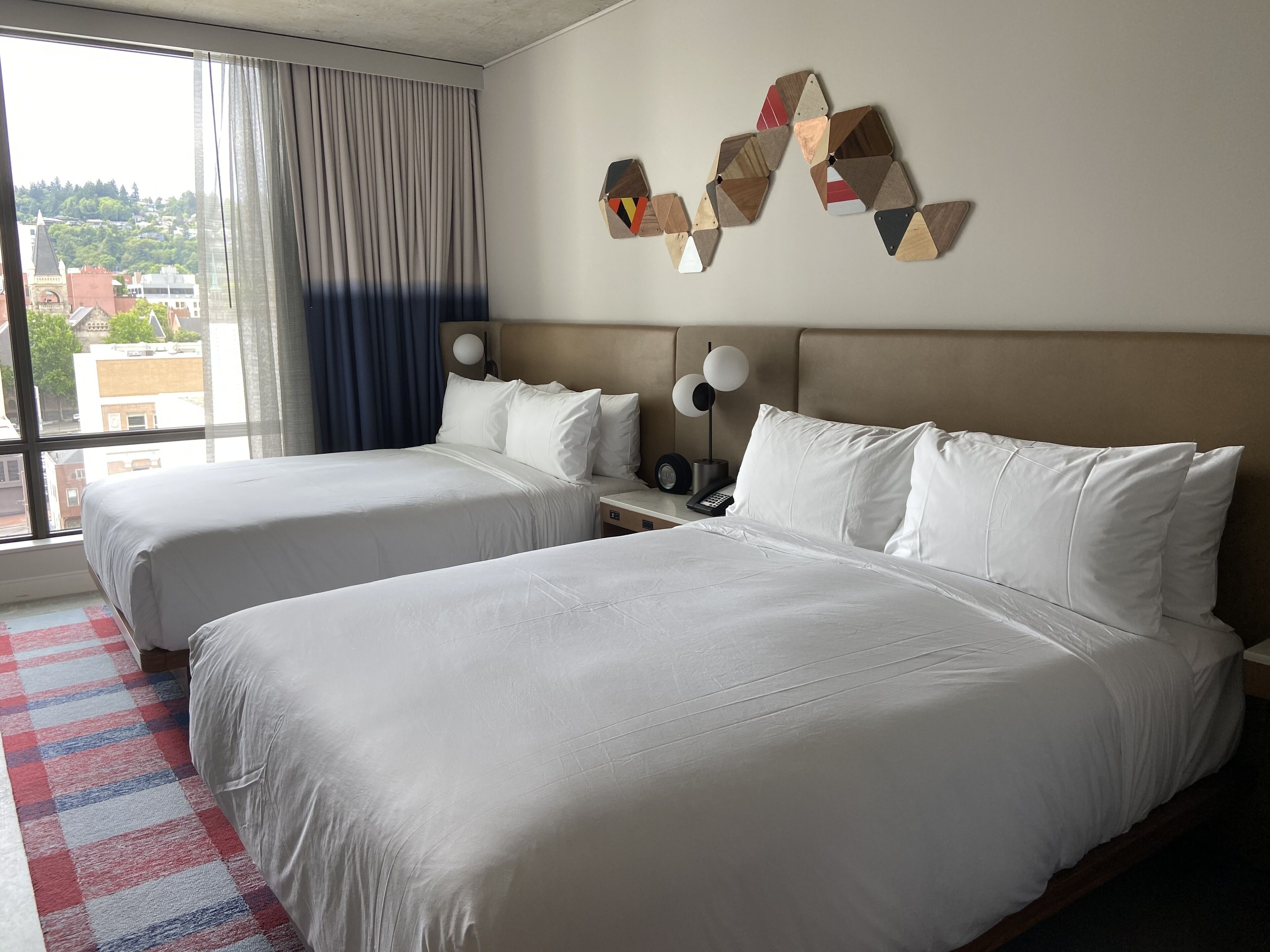 Quarto de hotel com duas camas, um tapete xadrez, uma obra de arte na parede e vista para prédios pela janela.