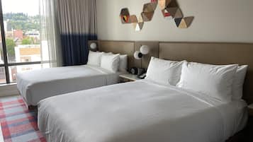 Quarto de hotel com duas camas, um tapete xadrez, uma obra de arte na parede e vista para prédios pela janela.