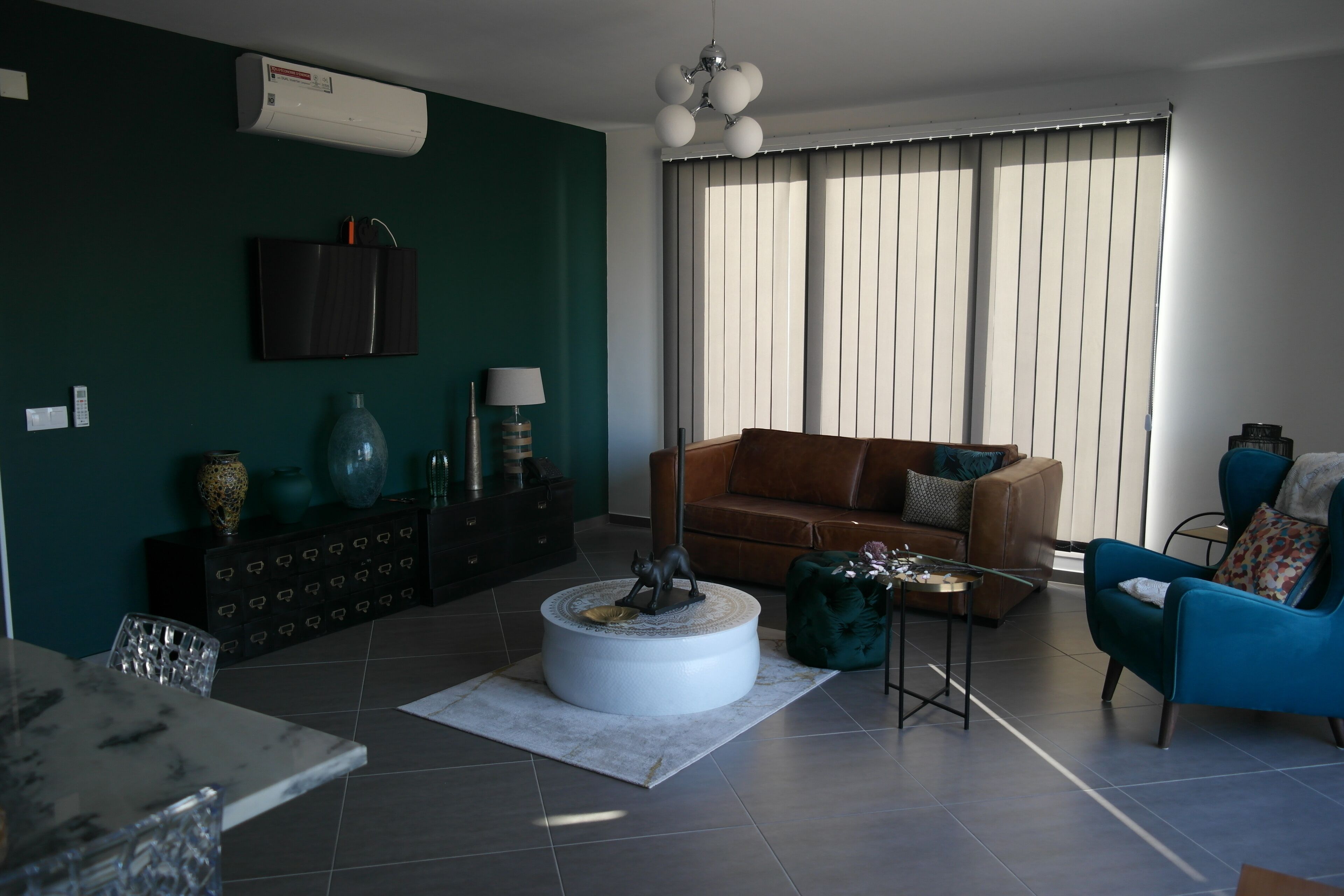 Uma sala de estar moderna com um sofá marrom, uma poltrona azul, uma mesa de centro redonda e uma TV fixada na parede.