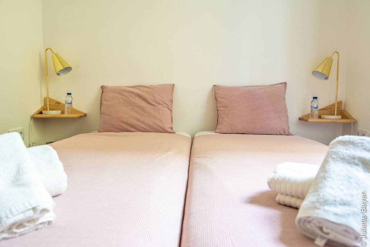 Quarto duplo com duas camas, cada uma com colcha rosa, duas almofadas iguais e duas mesas de cabeceira com luminárias e garrafas de água.