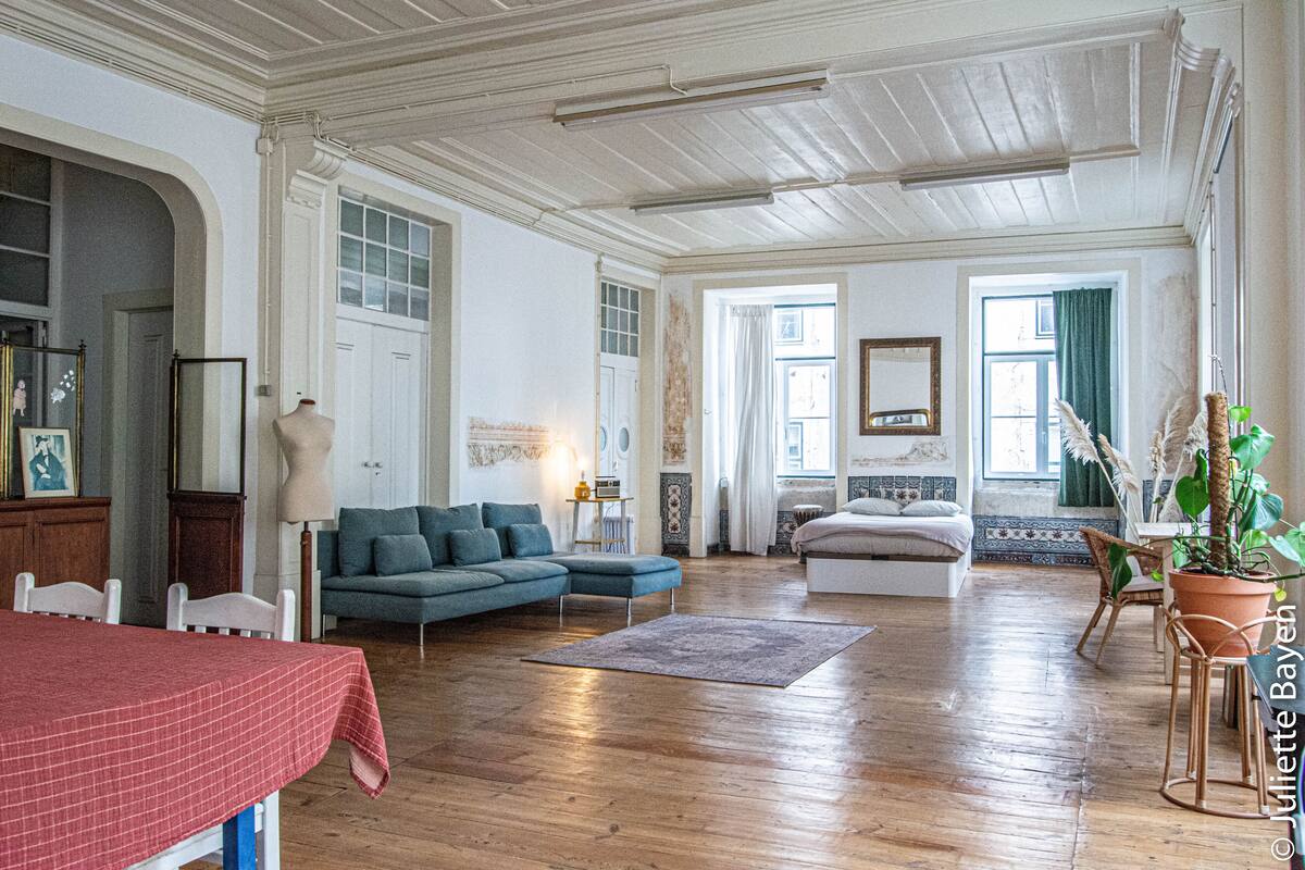 Uma sala de estar espaçosa com piso de madeira, um sofá azul, uma mesa de jantar com toalha vermelha e uma janela grande com cortinas.