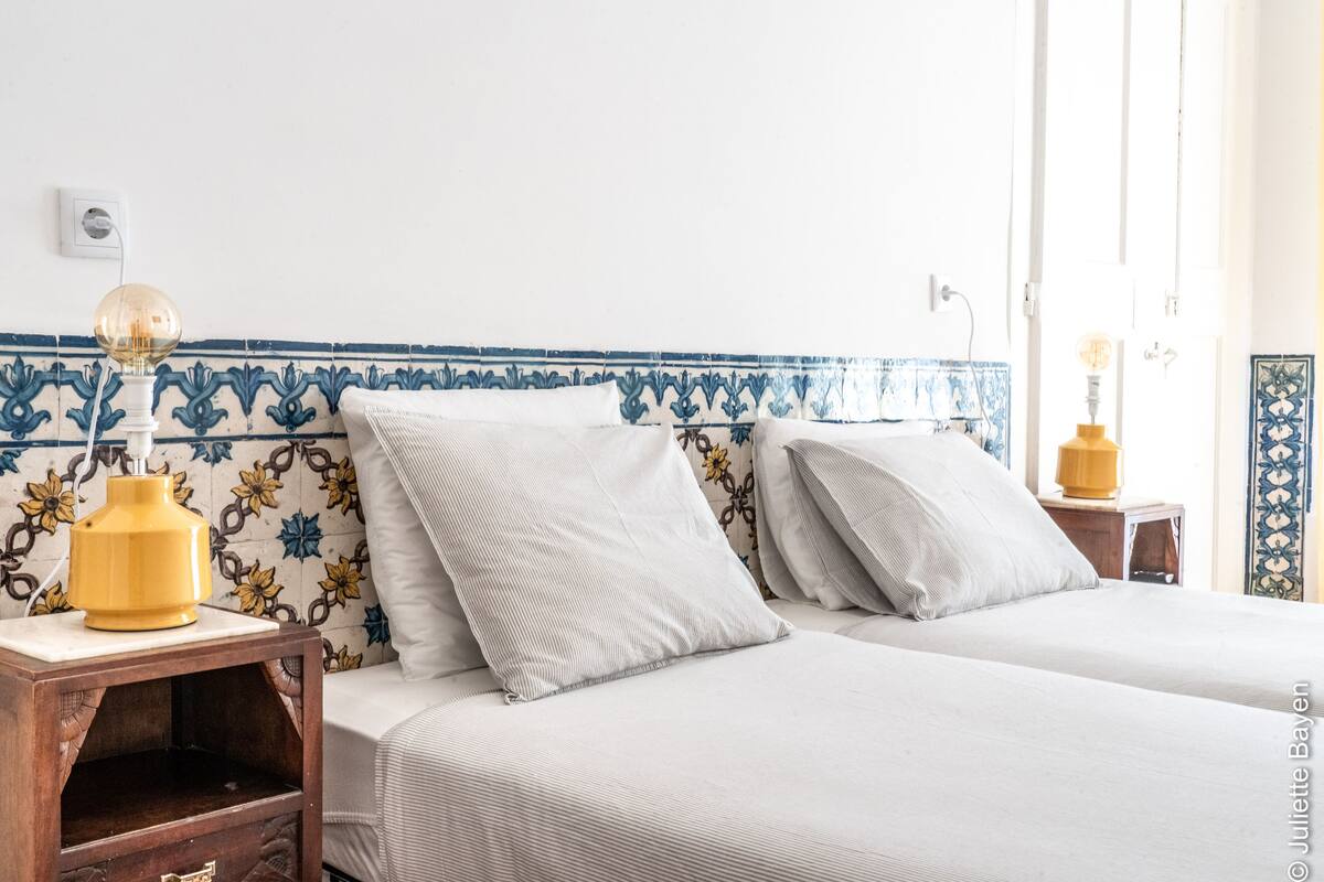 Quarto de hotel com duas camas, uma mesa de cabeceira de madeira com abajur e um painel de azulejos decorativo.