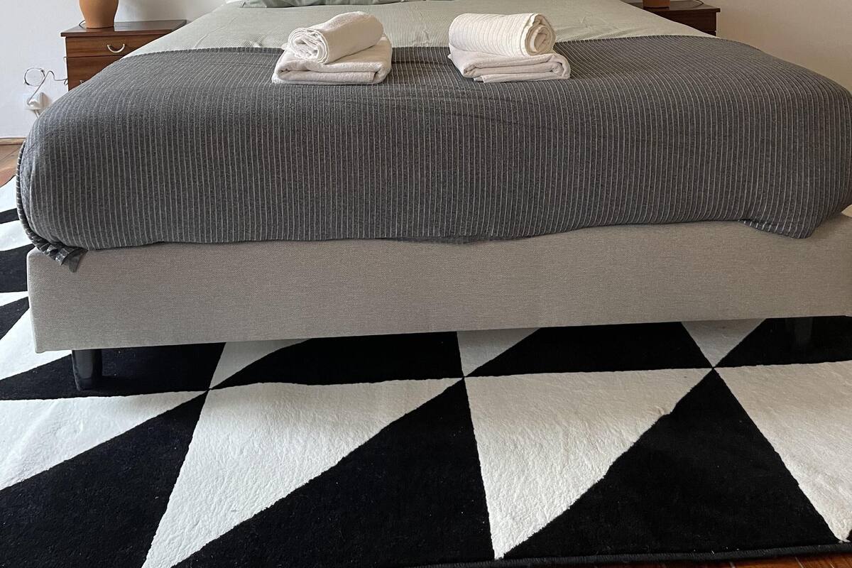 Cama bem arrumada com colcha listrada em cinza e branco, dois travesseiros e duas toalhas enroladas sobre a cama. Há duas mesas de cabeceira com abajures, e duas fotos emolduradas na parede.