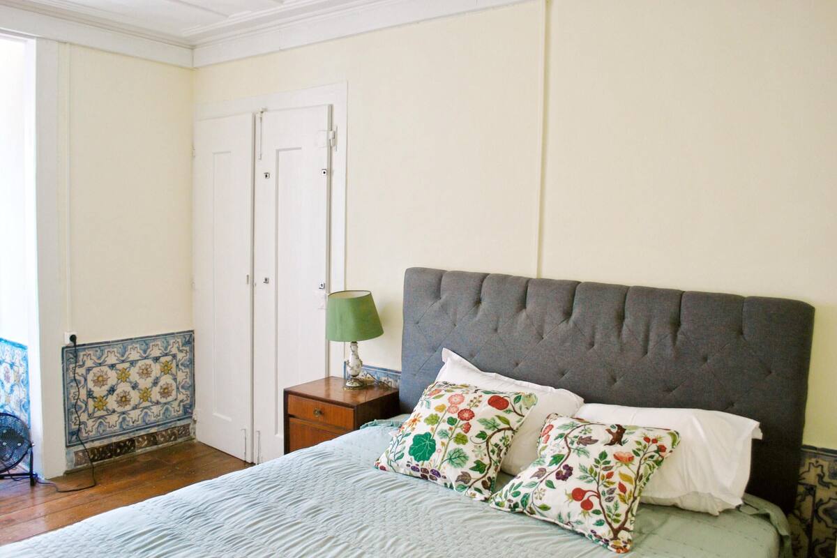 Quarto com cama, mesa de cabeceira, abajur e porta.