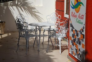 Área de estar externa com metal, incluindo mesa e cadeiras, uma placa com a inscrição 'Refugio Hostel Fortaleza' e uma parede vermelha com padrão geométrico.
