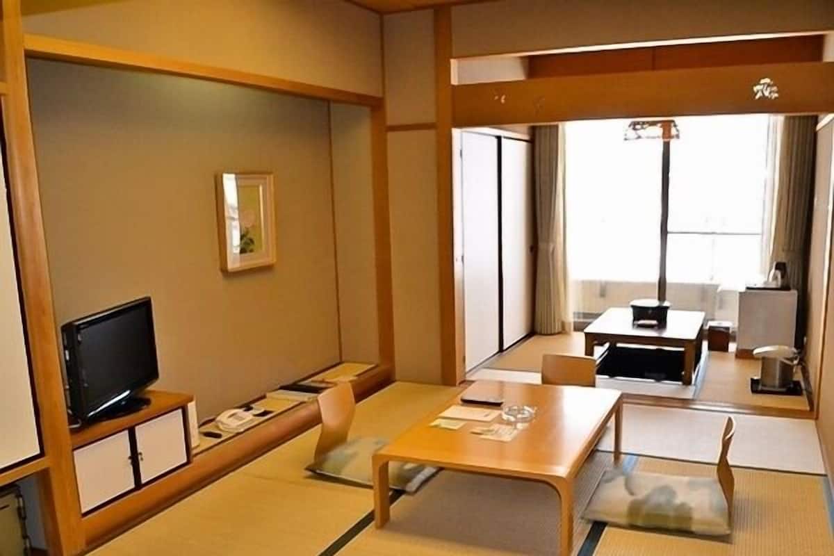 Um quarto tradicional em estilo japonês, com tatames, uma mesa baixa, cadeiras, televisão e portas deslizantes que dão para outro cômodo.
