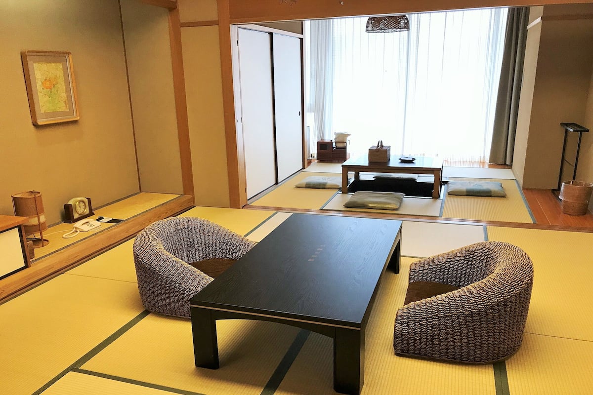 Um quarto japonês tradicional com tatames, uma mesa baixa, duas áreas de estar acolchoadas e uma porta de correr para uma varanda.